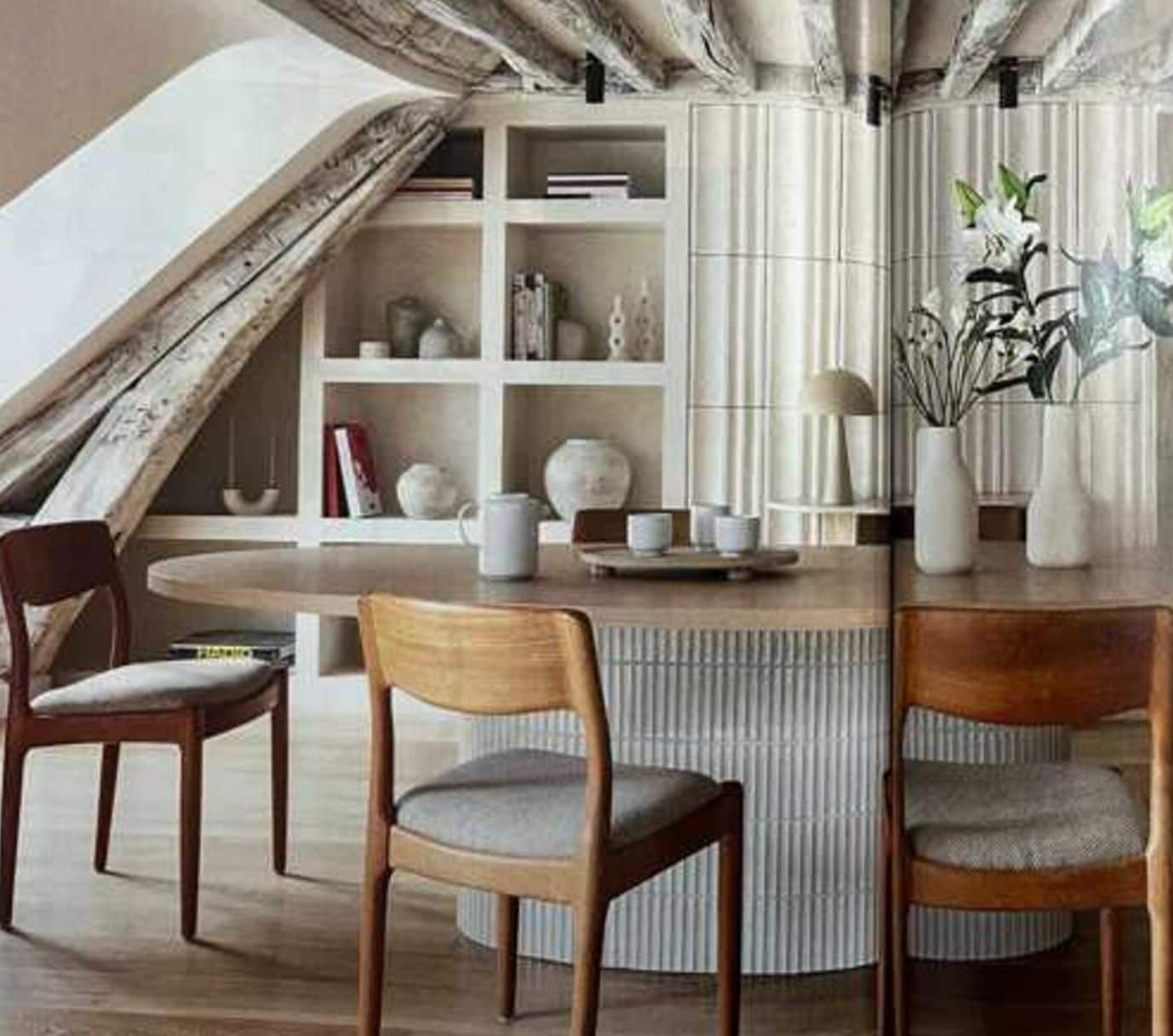 Uma área de refeições com uma mesa redonda, duas cadeiras de madeira com almofadas, uma estante com diversos objetos e uma planta decorativa.