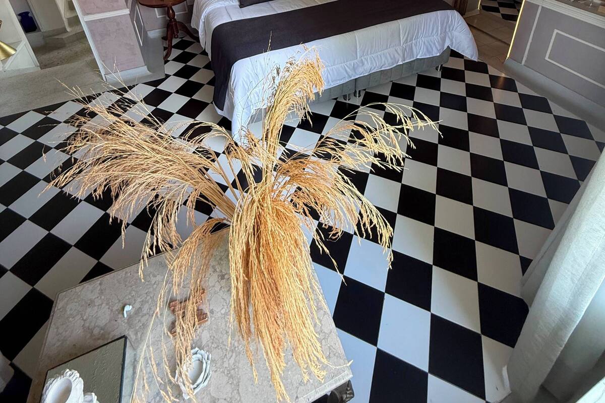 Quarto de hotel com chão xadrez, cama com roupa de cama branca, mesa redonda com um vaso e um espelho na parede.