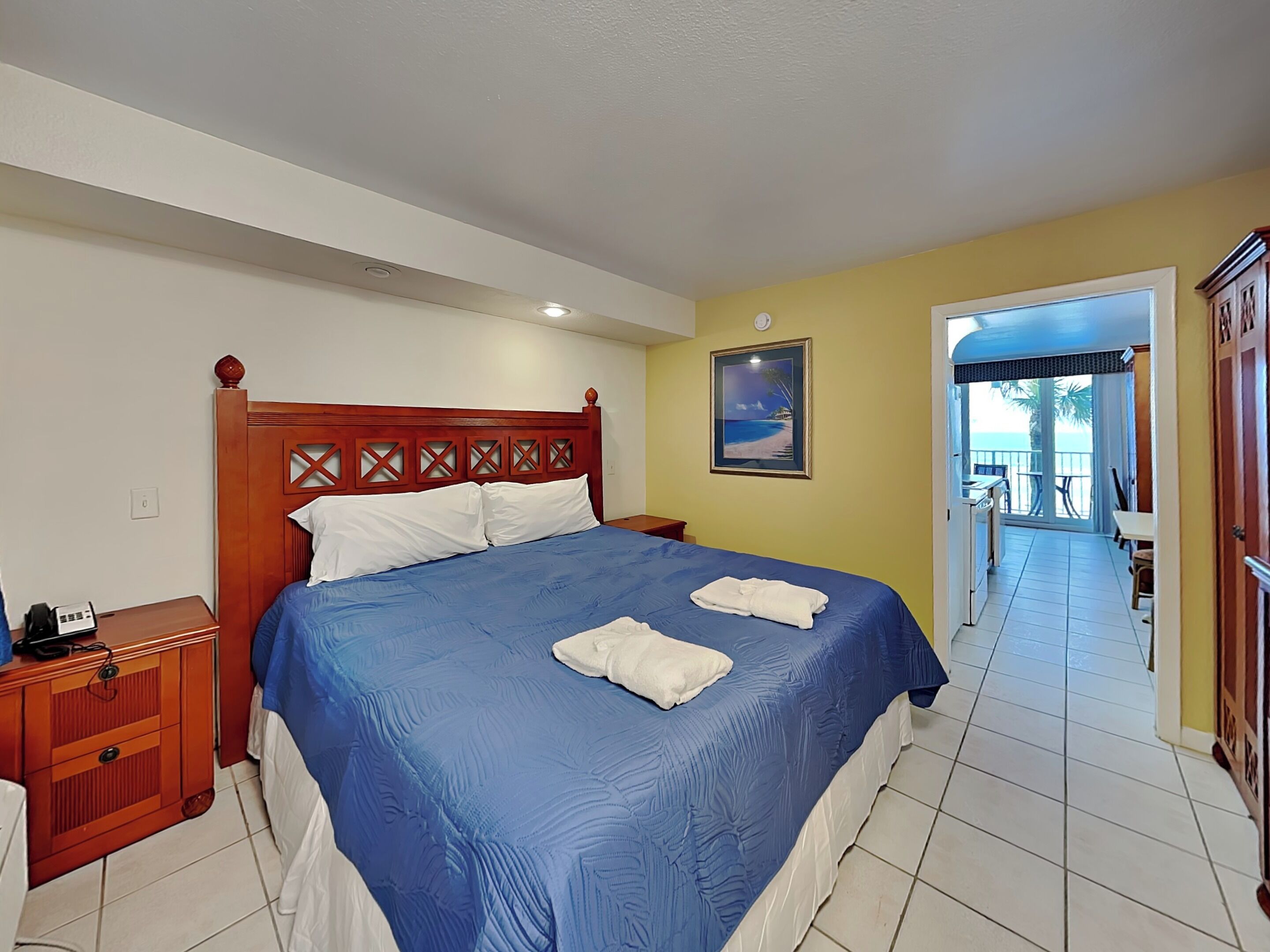 Quarto de hotel com uma cama grande, cabeceira de madeira, um quadro na parede e vista para a varanda externa.