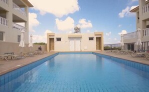 Piscina com superfície de água azul cristalina, rodeada por uma cerca baixa. Há espreguiçadeiras e guarda-sóis na área da piscina.