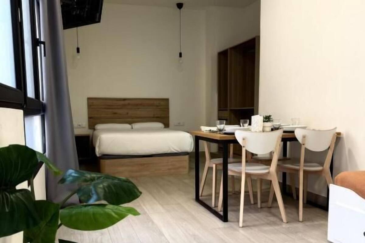 Quarto moderno com cama, mesa de jantar com cadeiras, televisão fixada na parede e um vaso de planta.