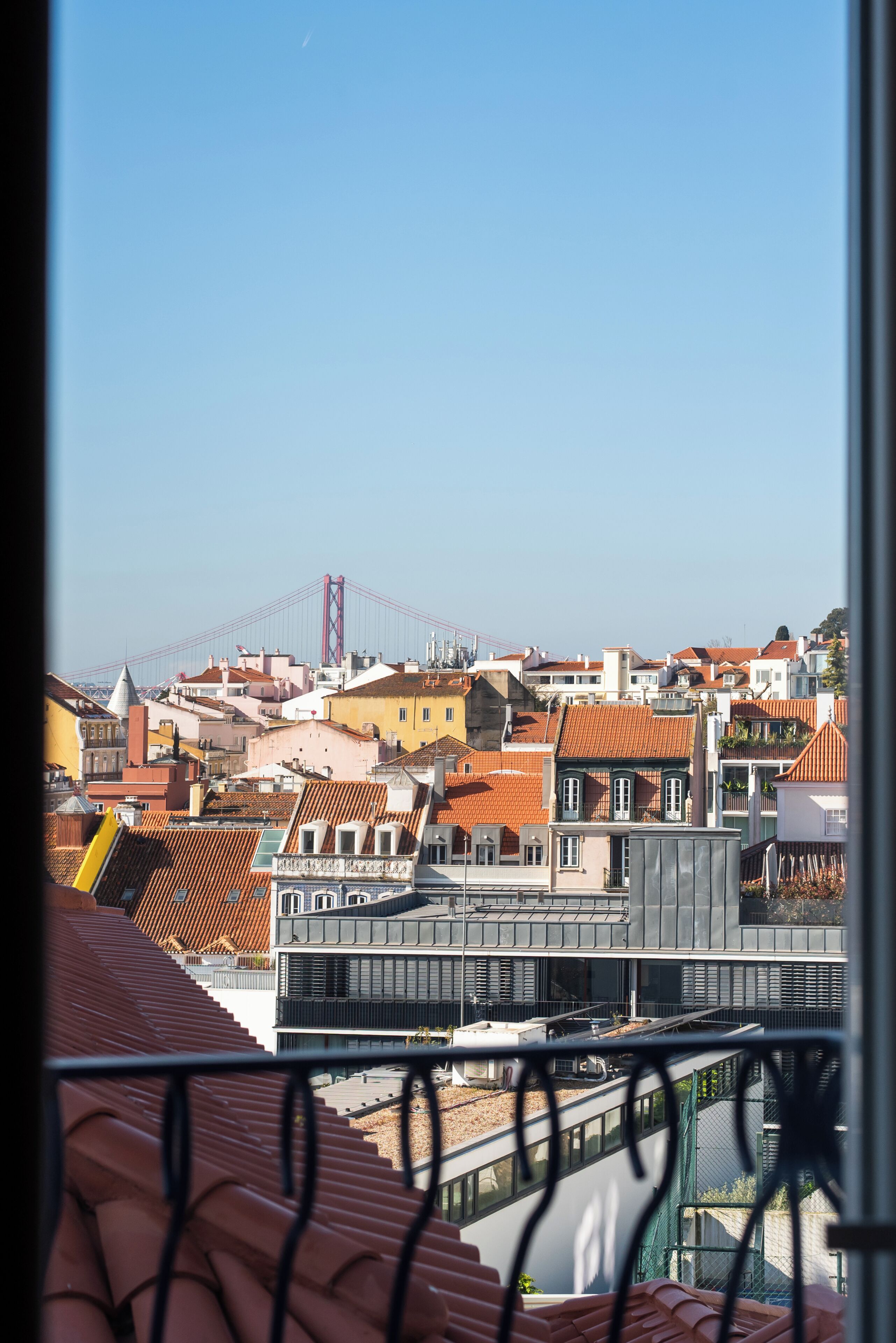 Paisagem urbana com uma ponte vermelha destacada ao longe, edifícios com telhados de terracota e uma construção moderna com telhado plano.