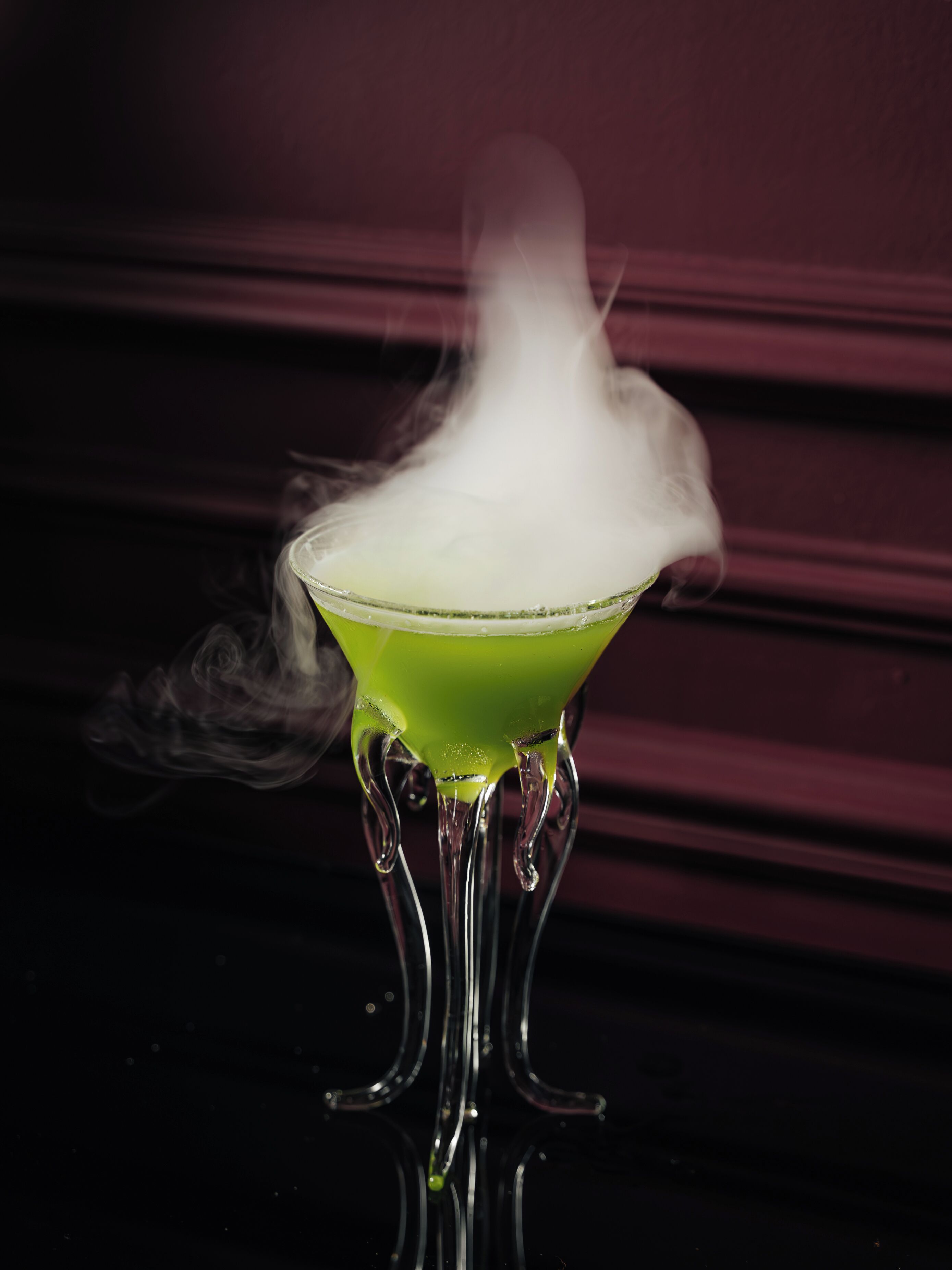 Um cocktail num copo de martini, com líquido verde e espuma branca, sobre um fundo escuro.