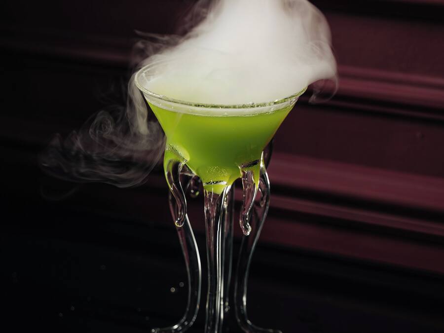 Um cocktail num copo de martini, com líquido verde e espuma branca, sobre um fundo escuro.