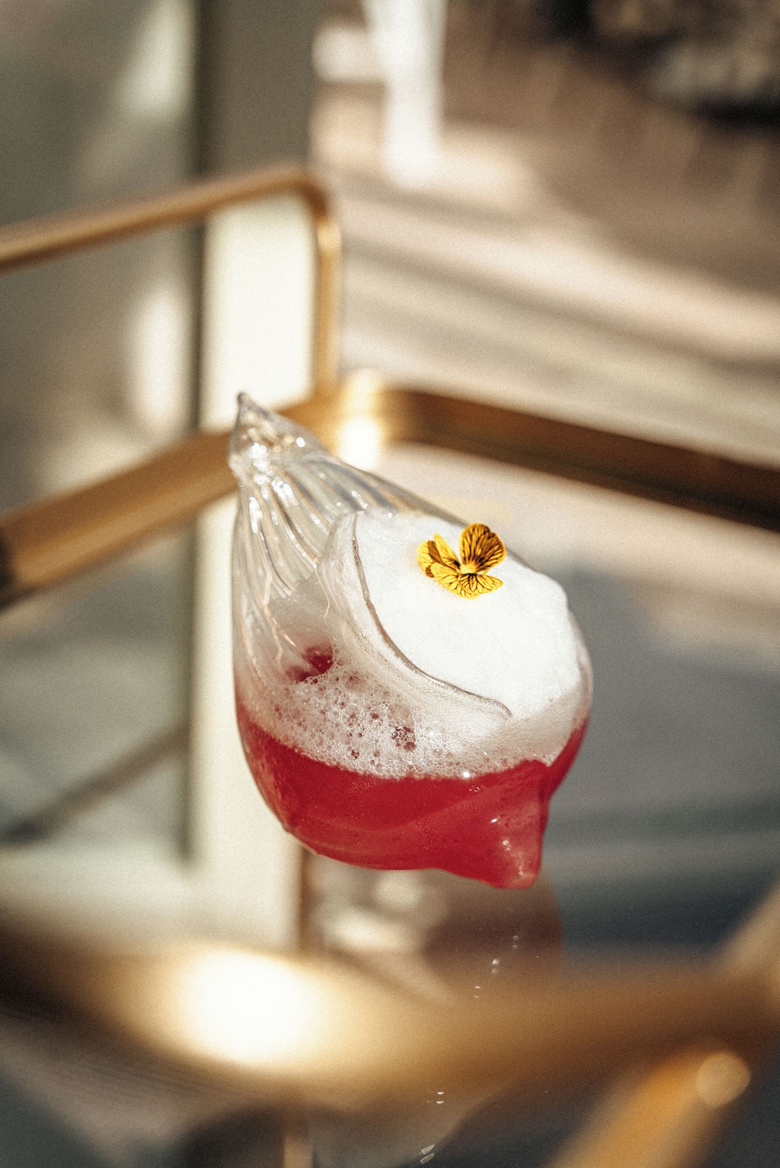Um copo de vidro transparente com líquido vermelho e espuma branca, apoiado em um suporte dourado.