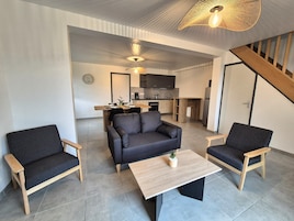 Uma sala de estar moderna com um sofá, duas poltronas e uma mesa de centro. Cozinha visível ao fundo.
