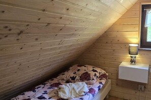 Quarto pequeno com teto inclinado, paredes de madeira, uma cama de solteiro com roupa de cama floral e uma luminária de cabeceira.