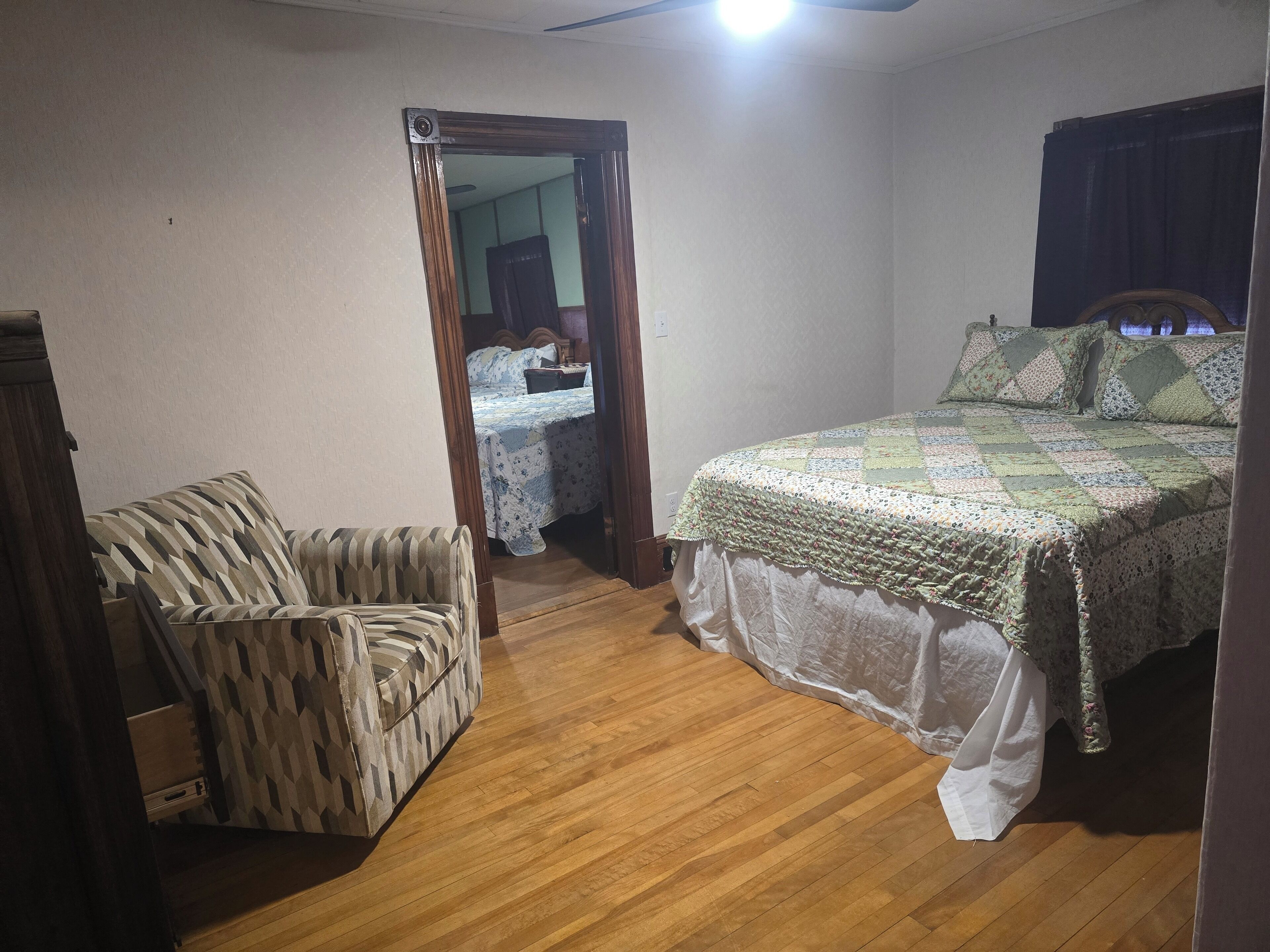Um quarto com piso de madeira, uma cama com colcha, uma cadeira e um guarda-roupa.
