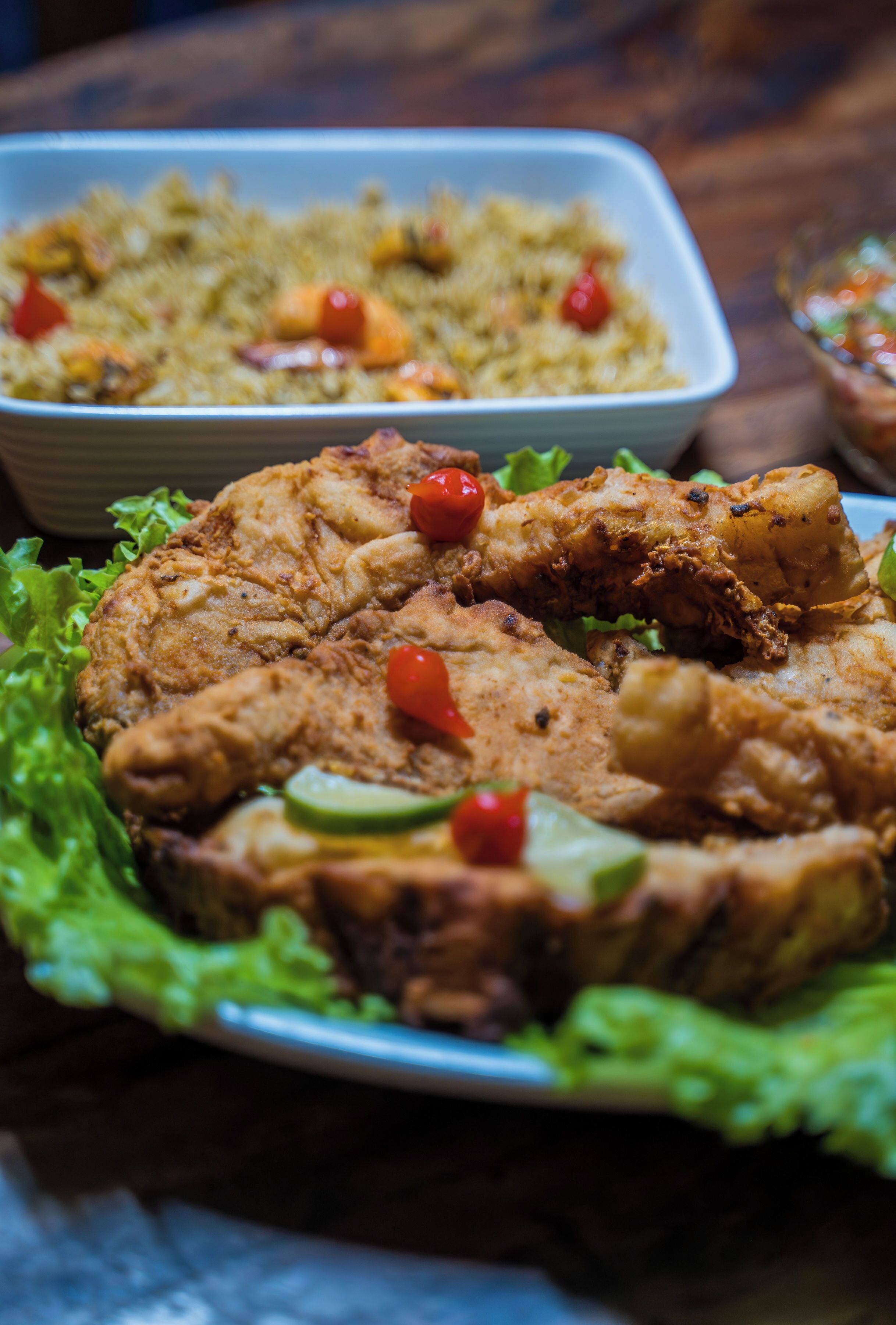 Um prato de frango frito com gomos de limão e tomates cereja, e uma tigela de arroz ao fundo.