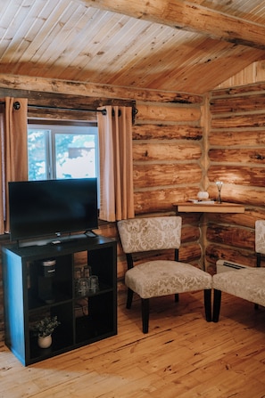 Interior de uma cabana de madeira com televisão sobre um suporte, duas poltronas estofadas, uma mesinha com um objeto decorativo e uma janela com cortinas.