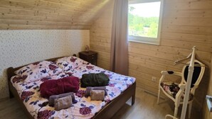 Quarto com teto de madeira, cama com roupa de cama florida, mesa de cabeceira, cadeira e janela com vista para vegetação.