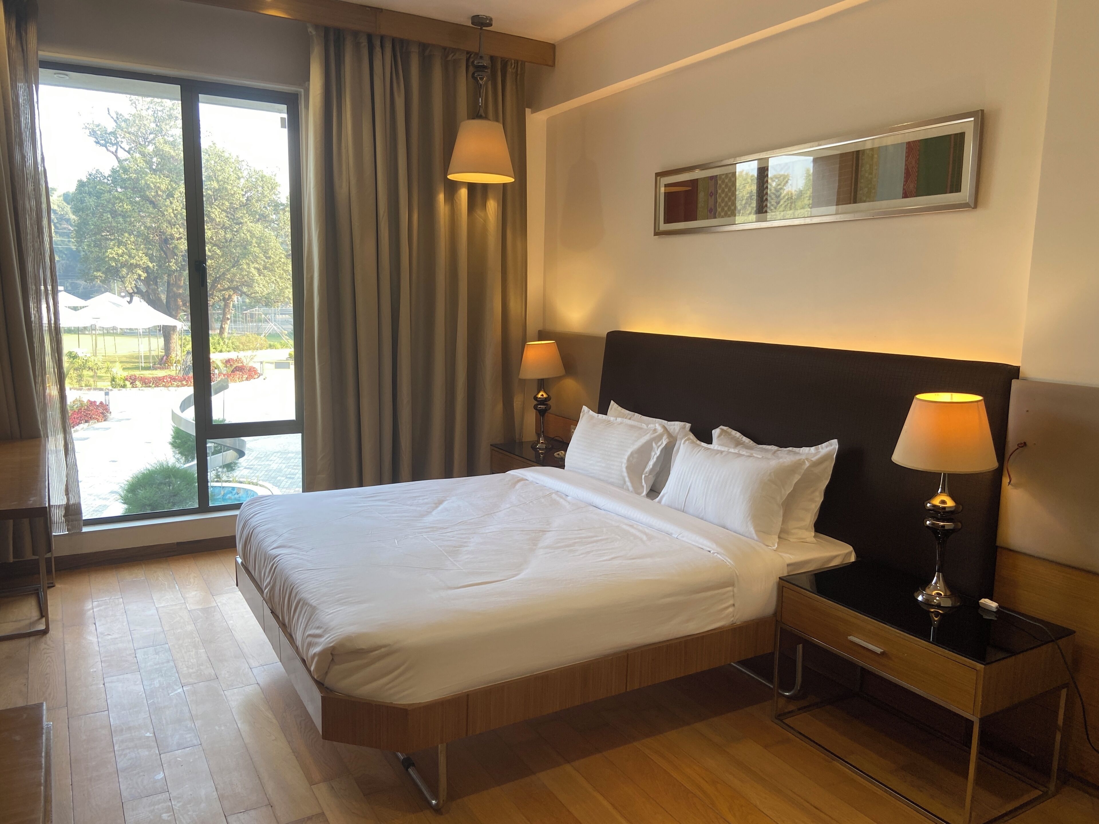 Quarto de hotel com uma cama grande, duas luminárias de cabeceira, um criado-mudo, piso de madeira e vista para o exterior por uma grande janela.