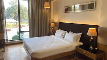 Quarto de hotel com uma cama grande, duas luminárias de cabeceira, um criado-mudo, piso de madeira e vista para o exterior por uma grande janela.