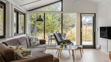 Uma sala de estar moderna com sofá, mesa redonda e cadeiras, janelas amplas e teto com vigas aparentes.