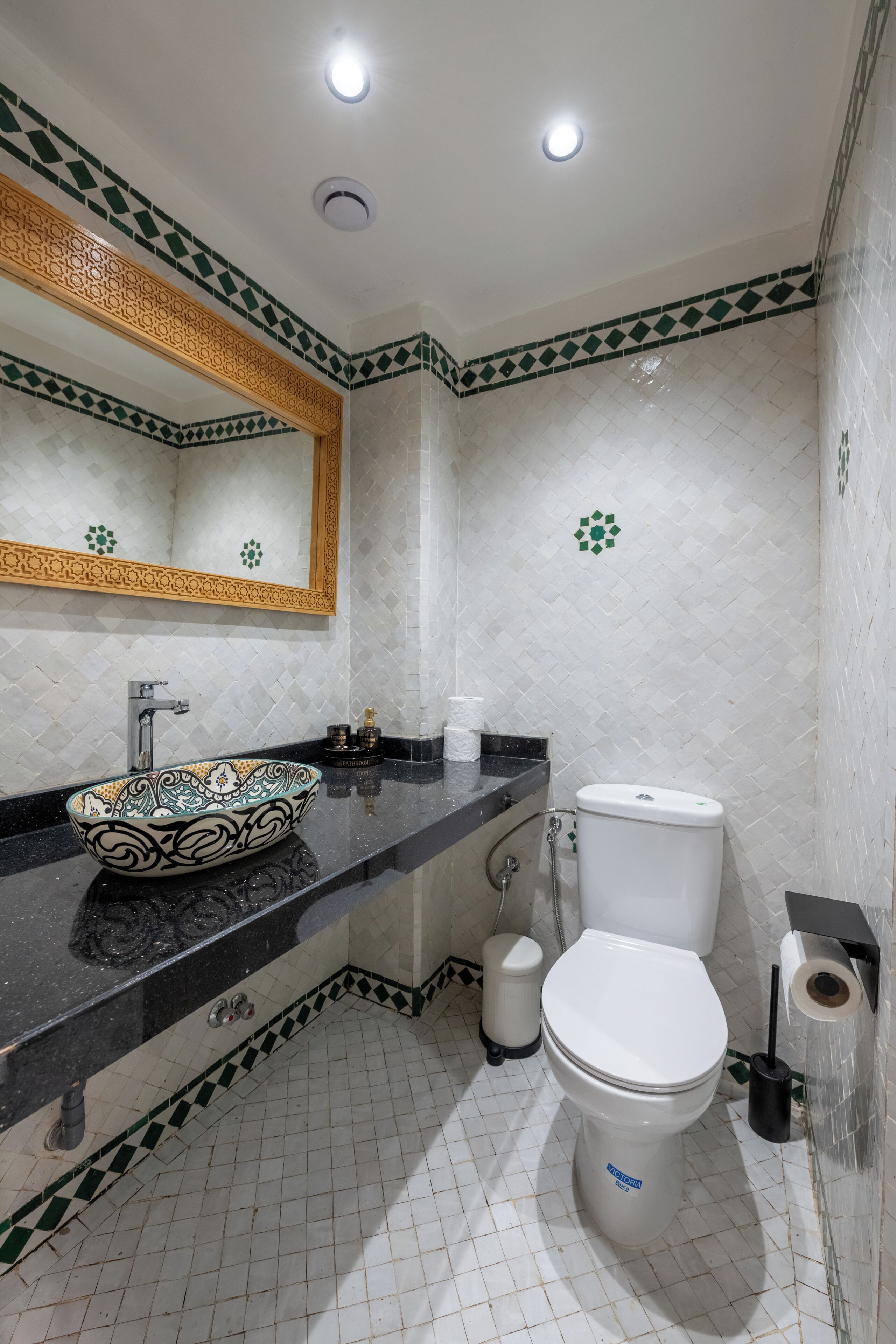 Um banheiro com piso de azulejos brancos, bancada preta, vaso sanitário branco e espelho com moldura decorativa.