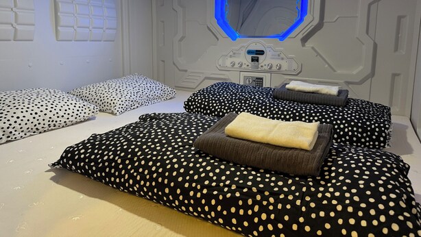Quarto com design futurista, apresentando um painel de controle central, um espelho hexagonal e roupa de cama com bolinhas.