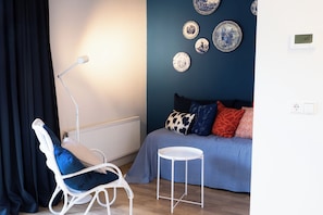 Um quarto com um sofá azul, uma cadeira branca, uma mesinha pequena branca e um abajur.