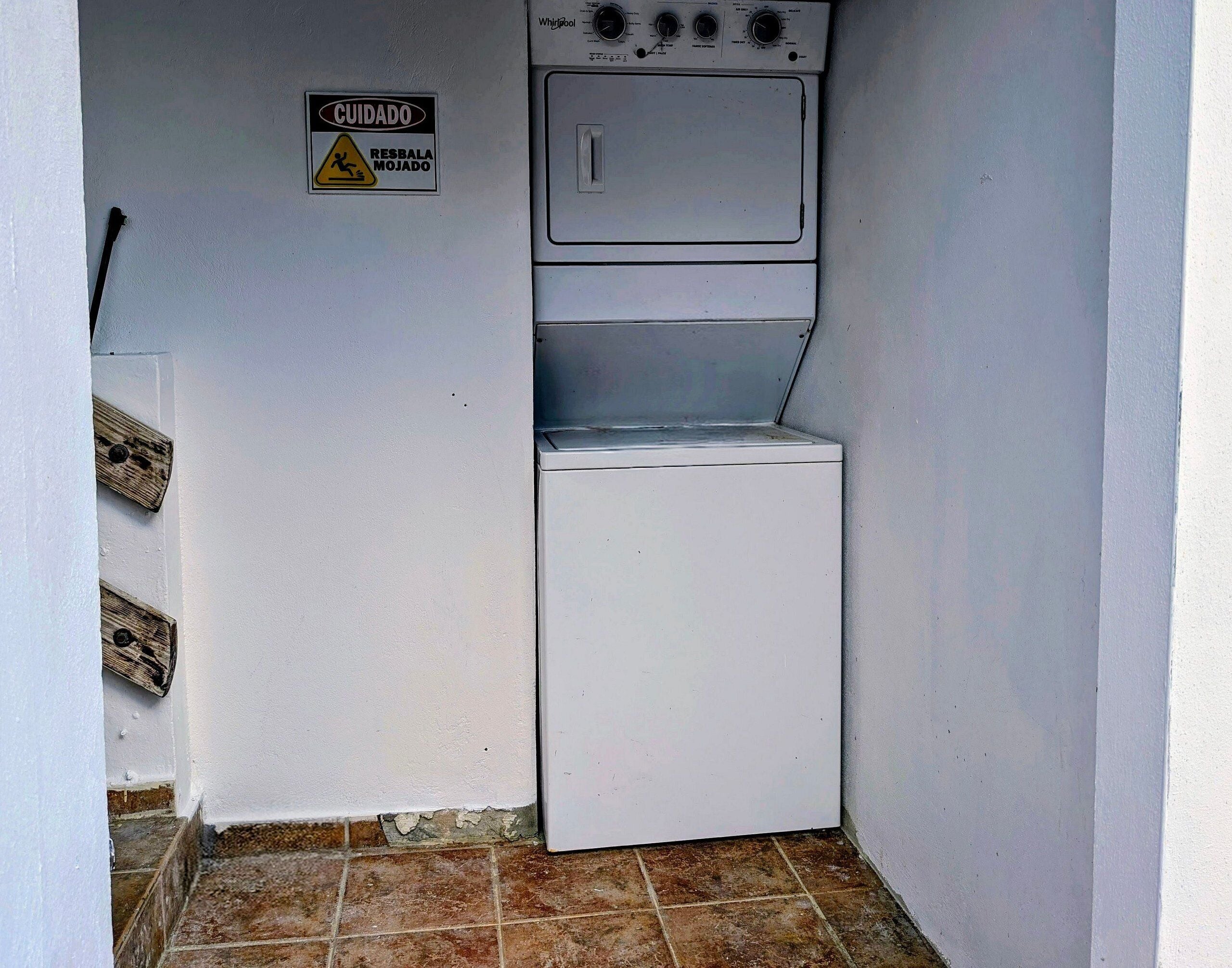 Uma lavanderia compacta e branca, com máquina de lavar e secadora Whirlpool, um aviso de segurança e piso revestido com azulejos.