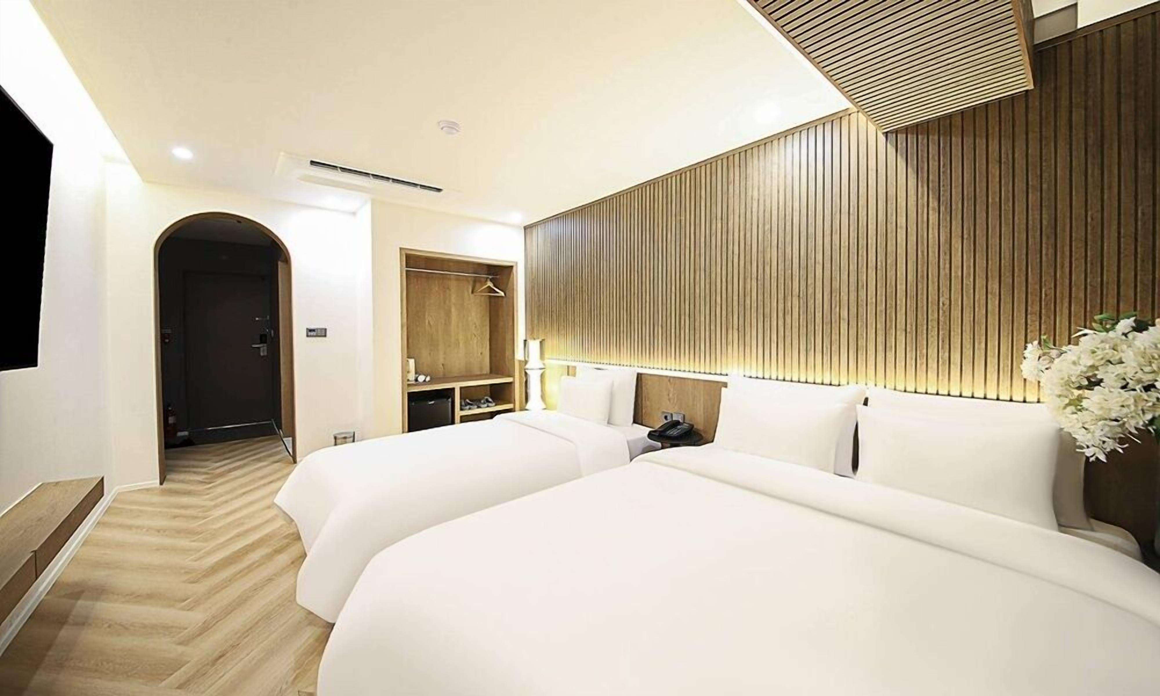 Quarto de hotel com duas camas, parede de painéis de madeira, televisão e corredor que dá para um armário.