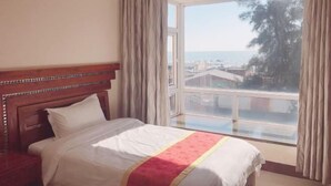 Quarto de hotel com uma cama grande, cabeceira de madeira, janela com vista para o mar e cortinas.