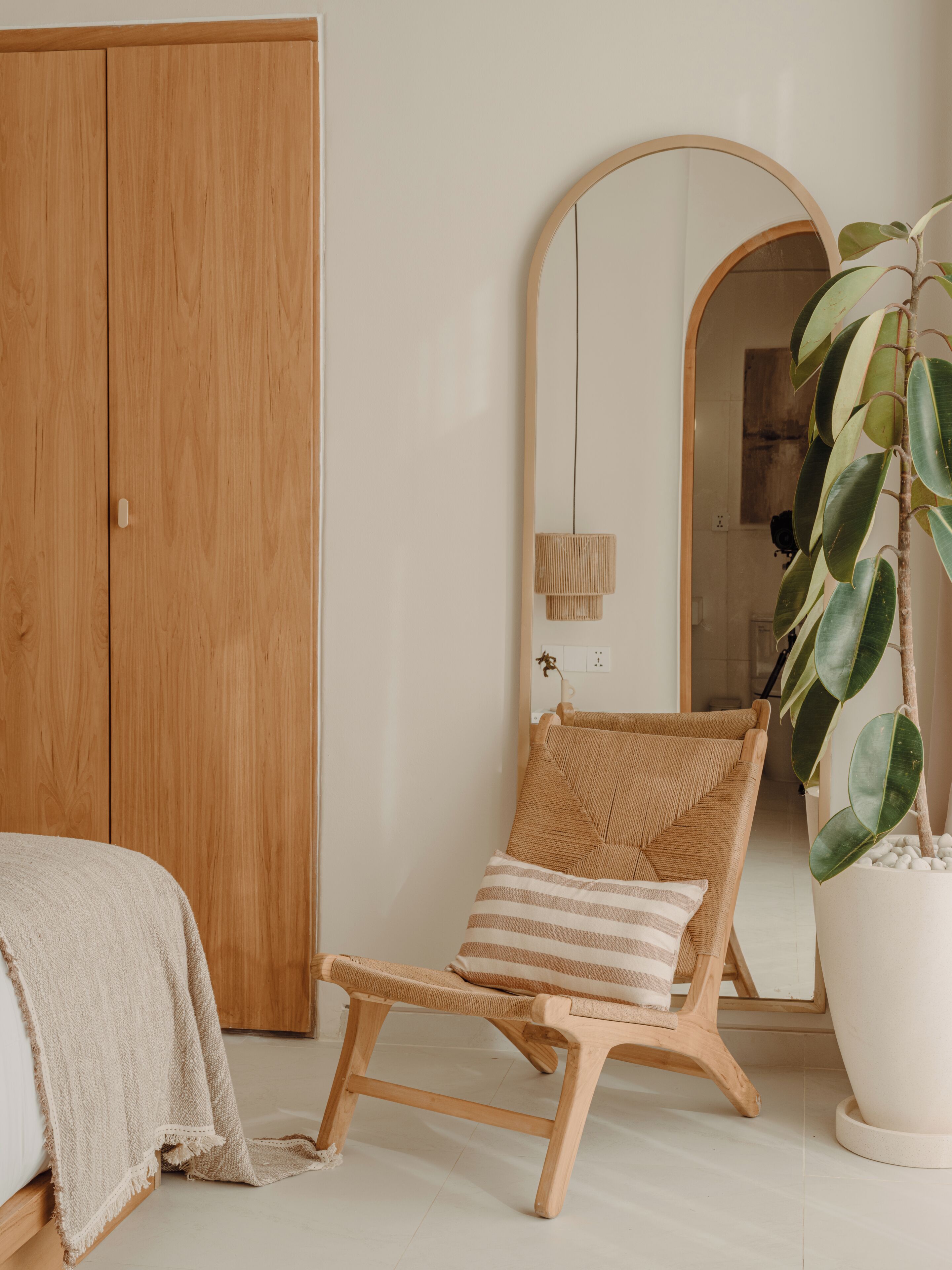 Quarto com guarda-roupa de madeira, cadeira de vime com almofada listrada, um espelho grande e uma planta em vaso.