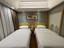 Quarto de hotel com duas camas, um quadro na parede, uma mesa de cabeceira com abajur e uma janela com persianas.