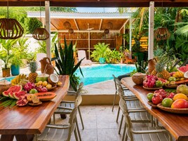 Uma área de refeições ao ar livre com piscina, cercada por plantas tropicais e diversas frutas sobre as mesas.