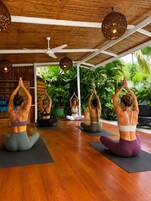 Aula de yoga em um espaço com piso de madeira, ventilador de teto e luminárias penduradas.