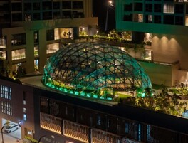 Um edifício moderno com uma grande estrutura de vidro em formato de cúpula no centro, rodeado por edifícios menores e vegetação.