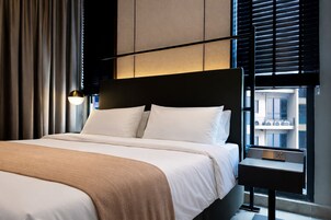 Quarto de hotel moderno com uma cama grande, mesa de cabeceira e janela com persianas.