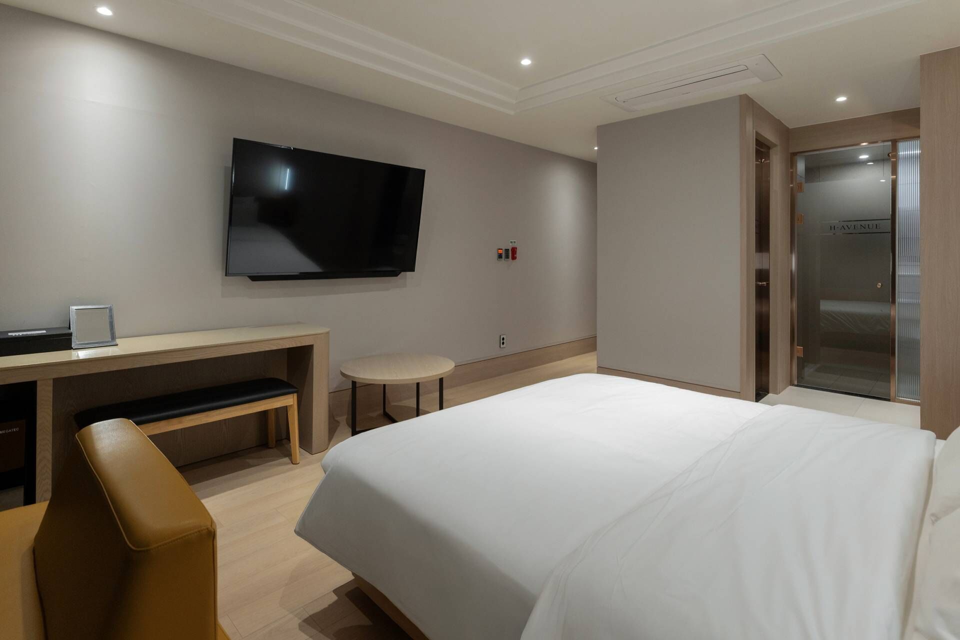 Quarto de hotel com cama, escrivaninha com cadeira, televisão e porta de vidro para um banheiro.