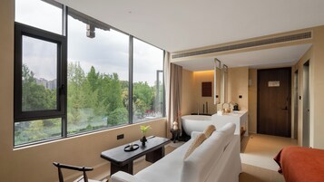 Quarto de hotel com uma janela ampla, um sofá, uma mesa pequena, um espelho, um banheiro com pia e porta-toalhas, e uma porta.