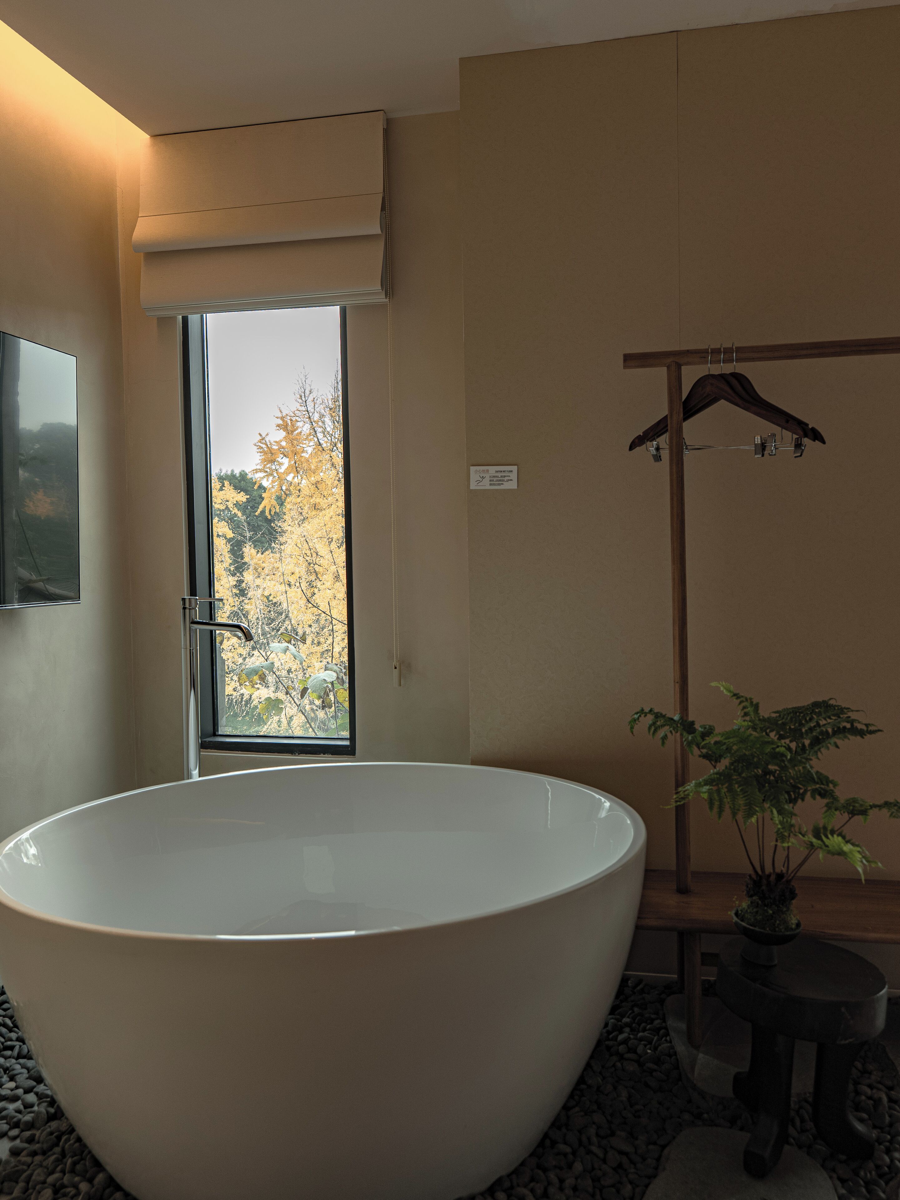 Um banheiro moderno com uma banheira grande e independente, uma janela com vista para árvores de outono e um suporte de madeira para cabides.