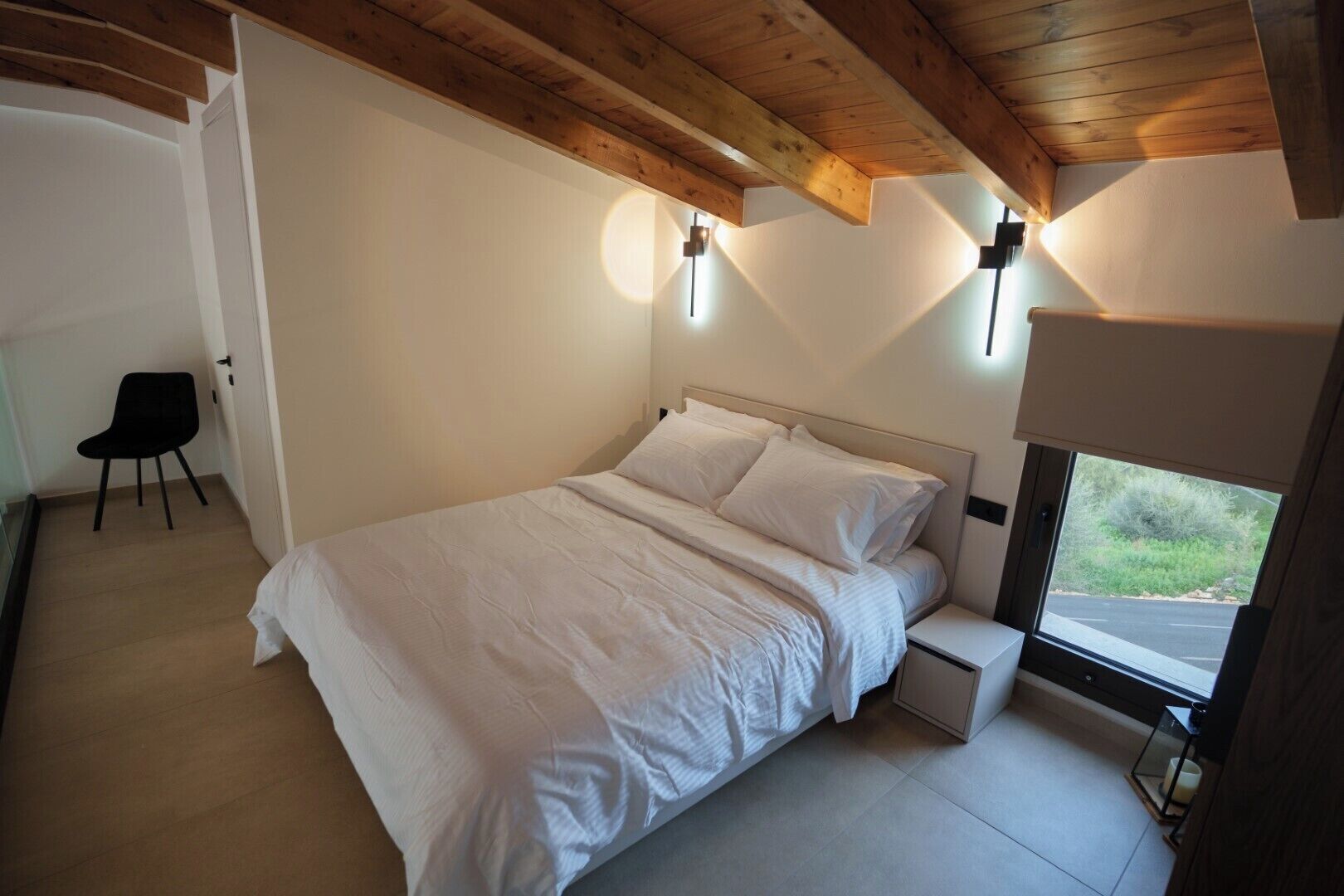 Quarto com cama, mesa de cabeceira, luminária, poltrona e janela com vista.