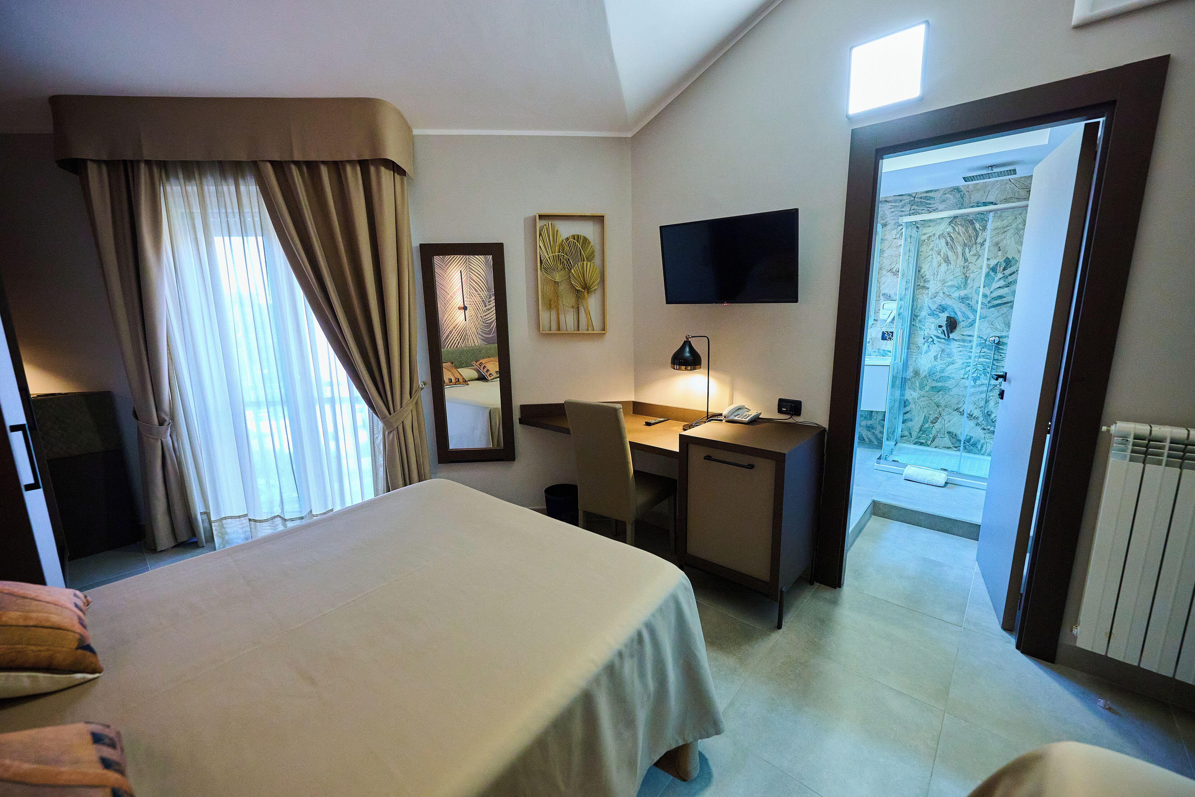 Quarto de hotel com cama, escrivaninha, cadeira, televisão e vista para uma varanda.