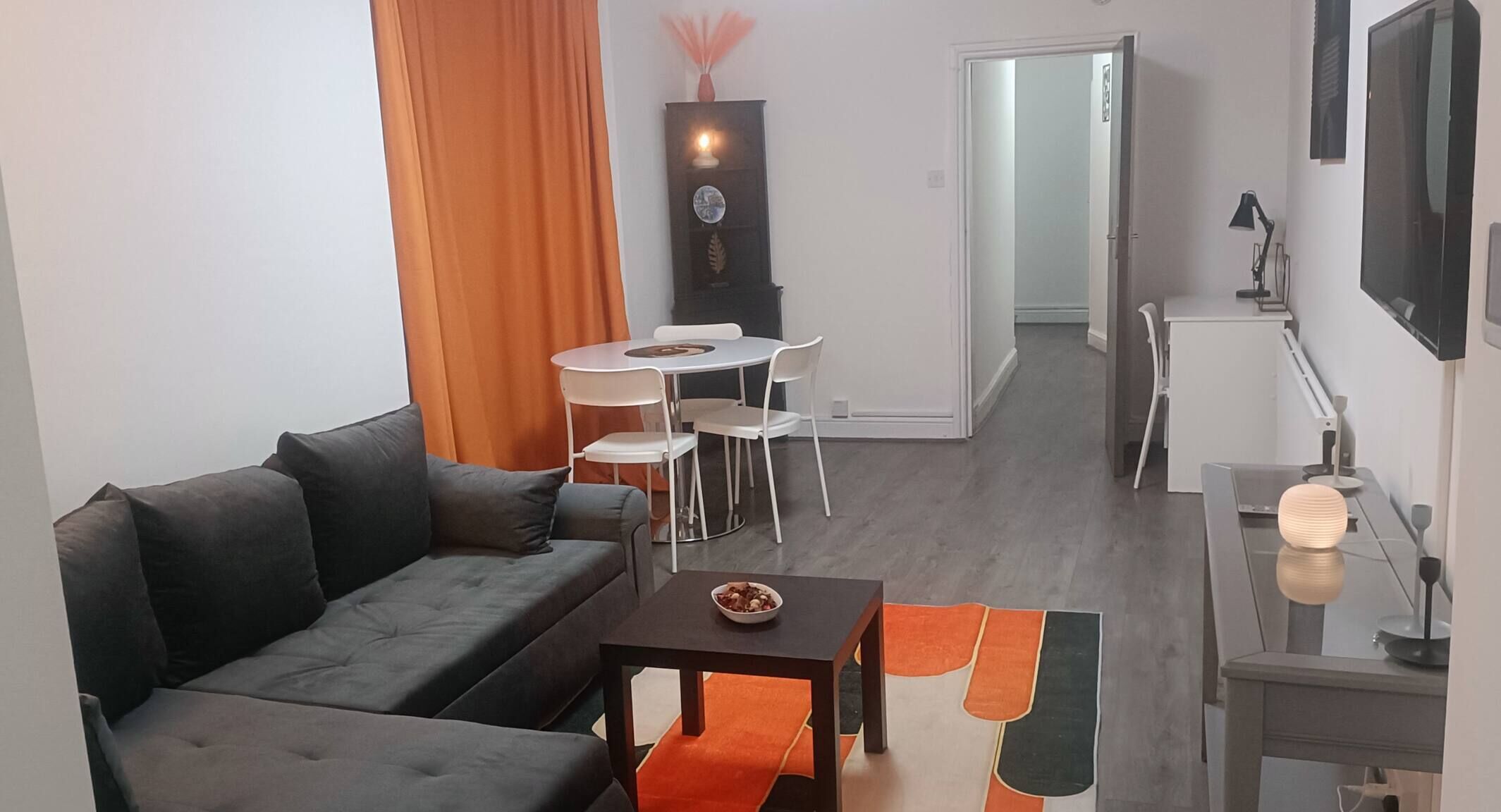 Uma sala de estar moderna com um sofá cinzento, uma mesinha com uma tigela de petiscos, uma mesa de jantar branca com cadeiras e uma TV na parede.