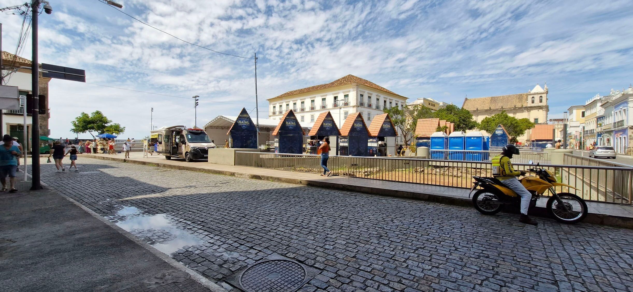 Uma rua de paralelepípedos com uma motocicleta amarela estacionada na lateral, um edifício branco com telhado vermelho ao fundo e um quiosque azul com a palavra 'NATAL' escrita nele.
