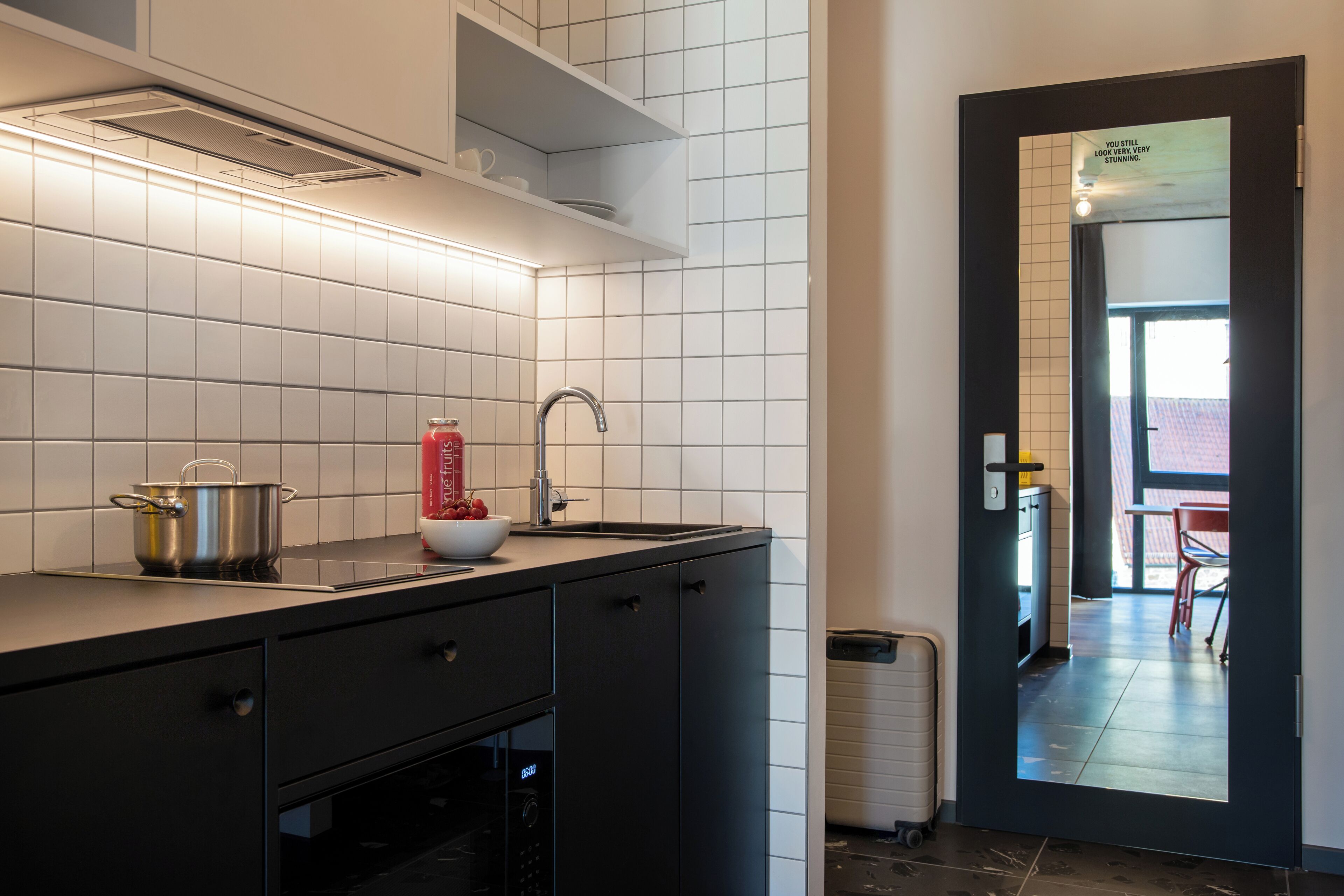Uma cozinha moderna com azulejos brancos, bancada preta, panela de aço inoxidável e um copo medidor vermelho.