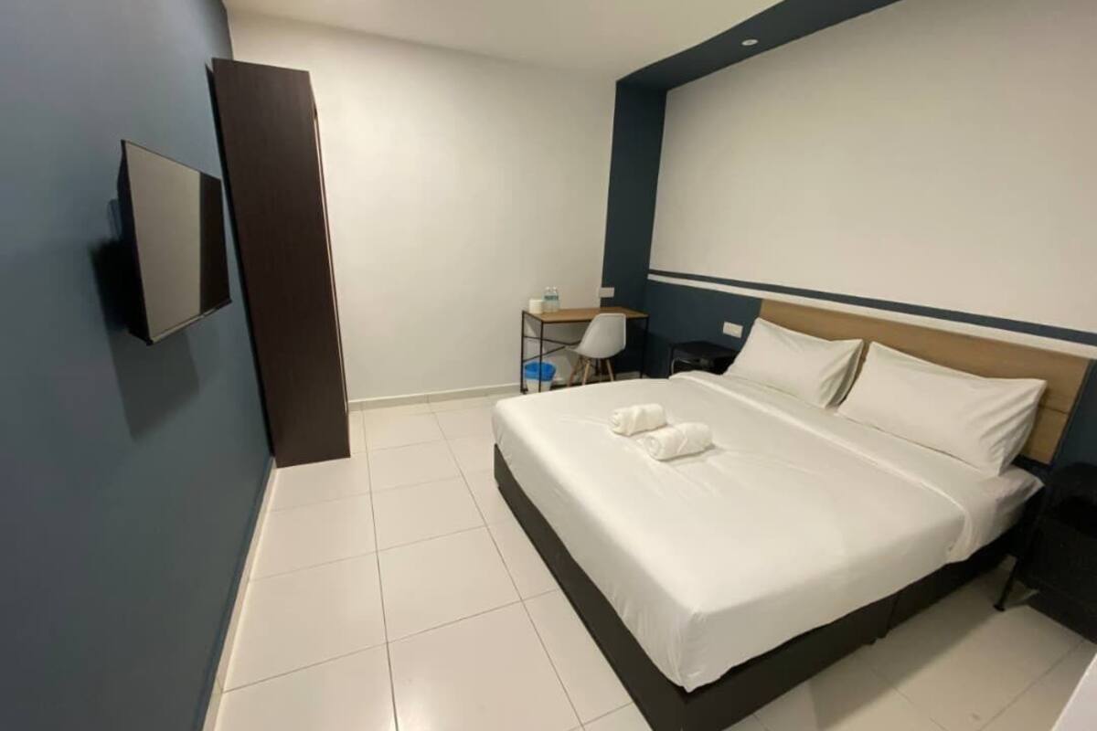 Quarto de hotel com cama, escrivaninha, cadeira, mesa de cabeceira e televisão na parede.