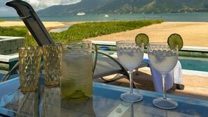 Uma mesa com dois copos de bebida transparente, uma garrafa de água e um copo de cocktail com um pedaço de limão, com vista para uma praia e montanhas.