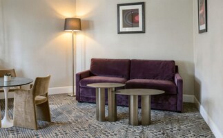 Quarto com sofá roxo, duas mesinhas redondas, um abajur e um quadro pendurado na parede.