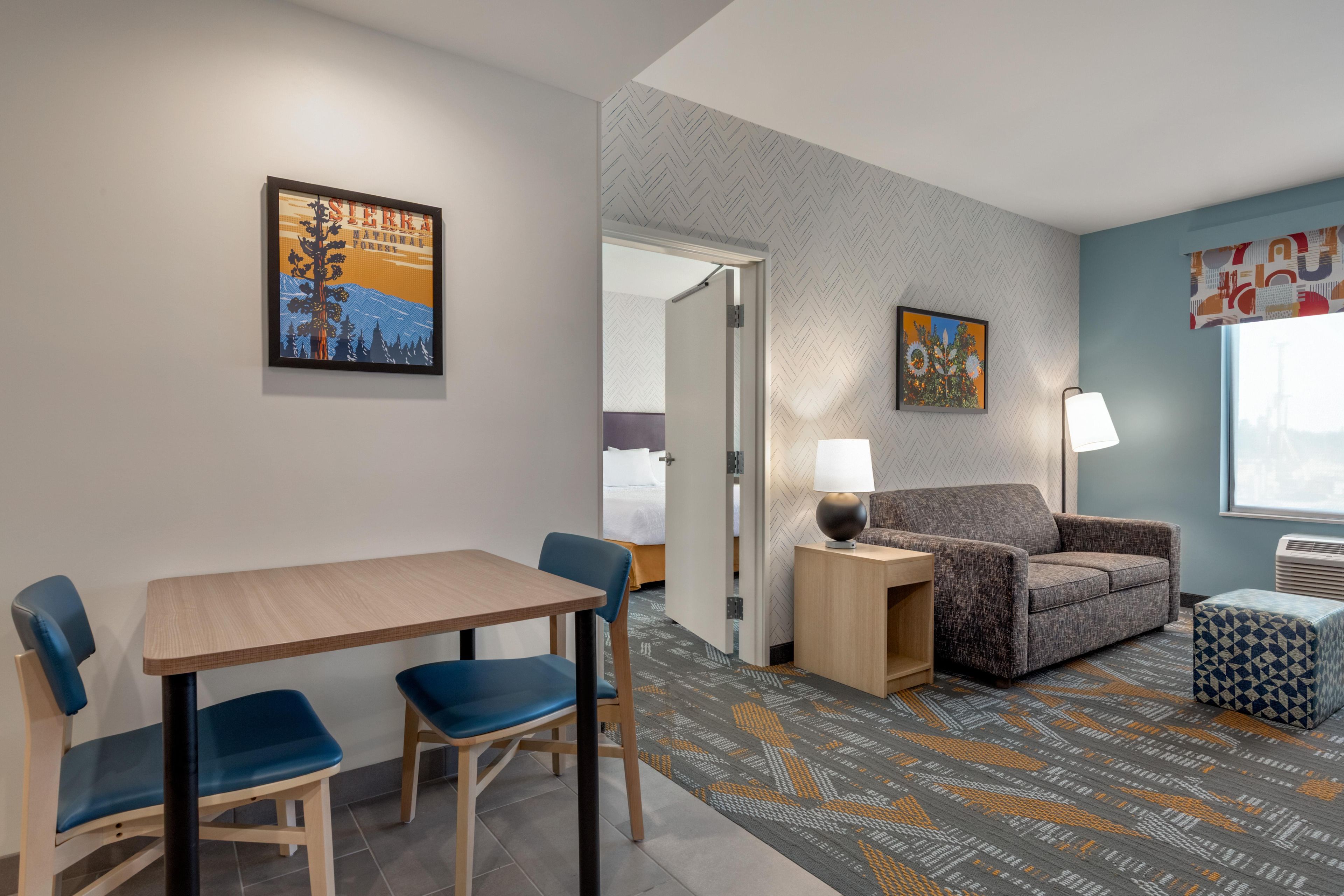 Quarto de hotel com mesa de jantar, duas cadeiras, um sofá, um abajur e um tapete estampado.