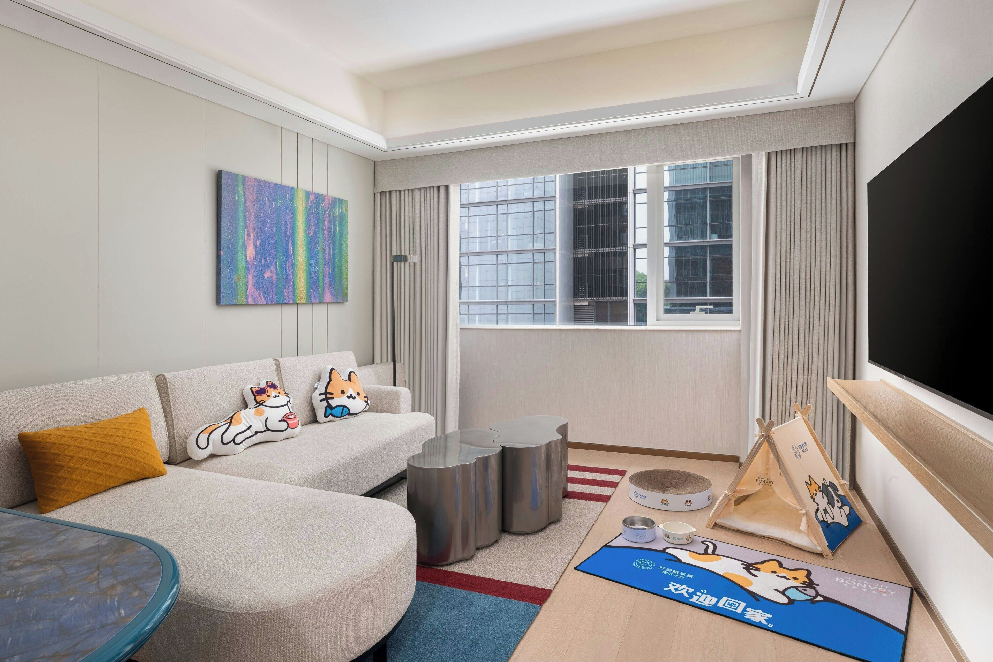Quarto de hotel moderno com sofá, uma mesa pequena, televisão, janela com cortinas e um quadro colorido na parede.