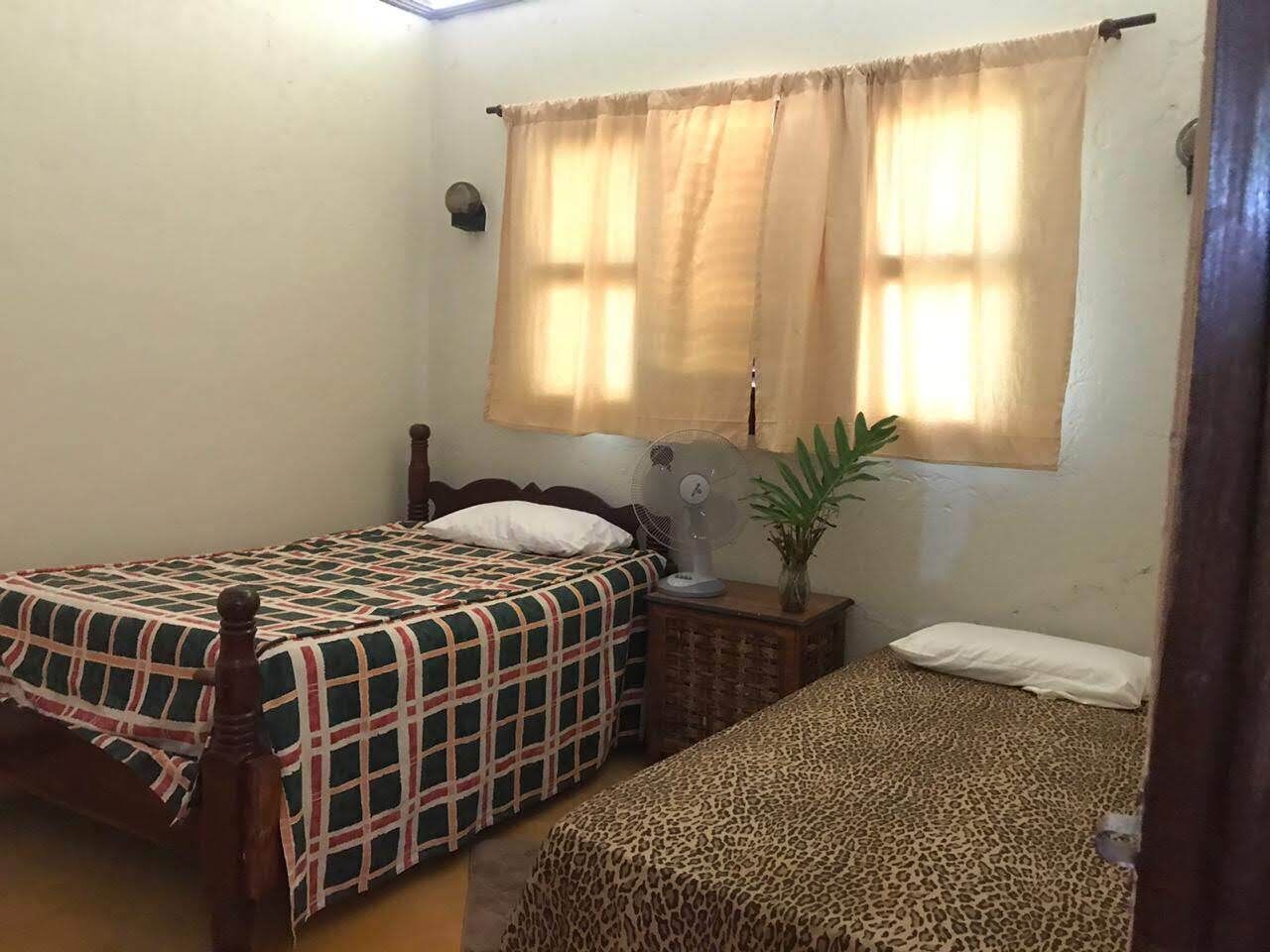 Quarto com duas camas, uma mesa de cabeceira com um ventilador, uma janela com cortinas e uma planta em vaso.