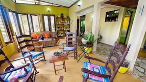 Uma sala de estar aconchegante com móveis de madeira, um sofá e uma mesa de centro. Há plantas em vasos e uma estante com diversos objetos.