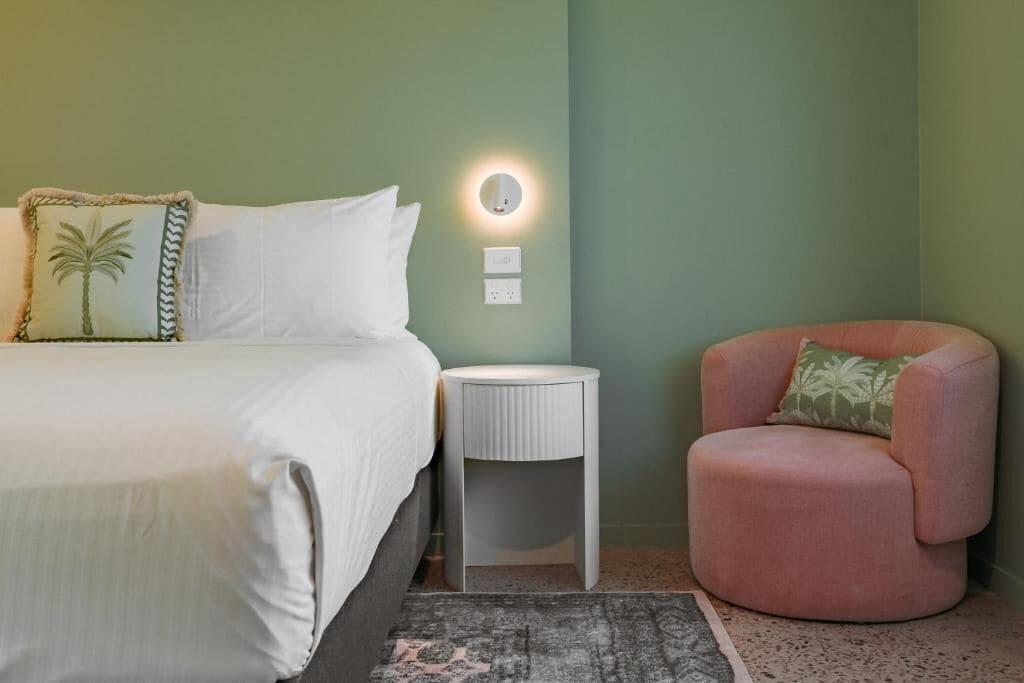 Quarto com cama, mesa de cabeceira, cadeira redonda e luminária na parede.