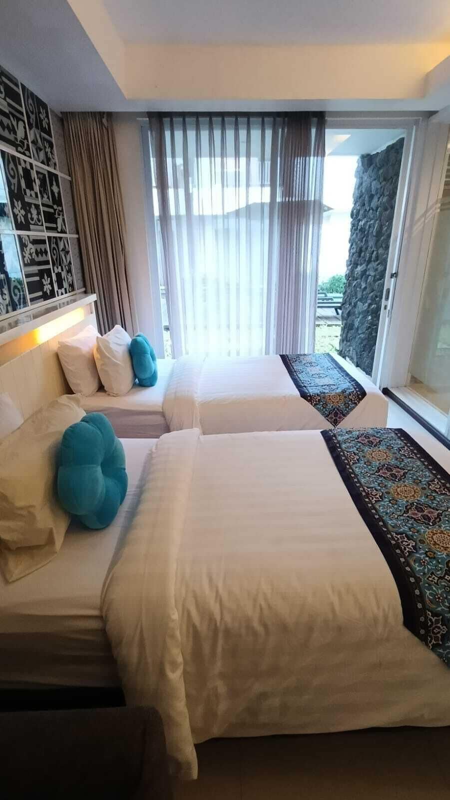 Quarto de hotel com duas camas, uma janela grande com cortinas, um tapete estampado e um detalhe decorativo na parede.