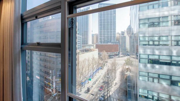 Vista pela janela com moldura metálica, mostrando um horizonte urbano com edifícios altos e uma rua com árvores e veículos.