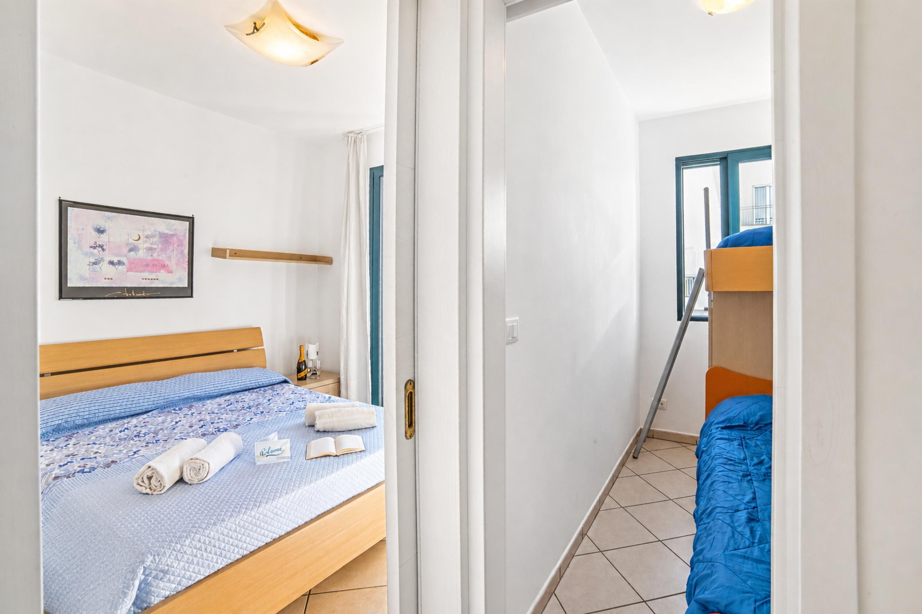 Quarto de hotel com cama, mesa de cabeceira, quadro emoldurado e vista através de uma porta aberta.