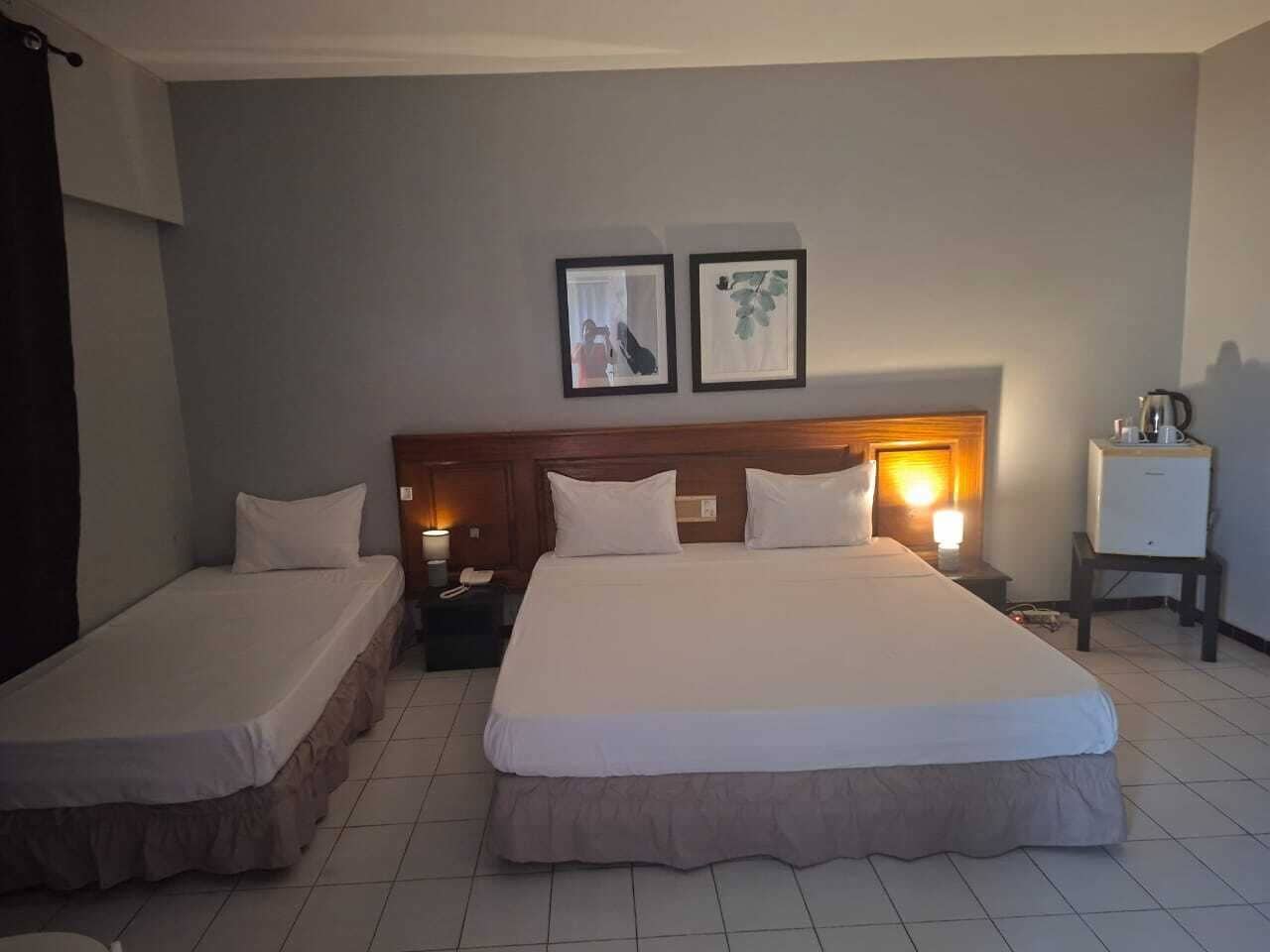 Quarto de hotel com duas camas de solteiro e uma cama de casal, cabeceira de madeira, abajures, uma pequena mesa com luminária e um refrigerador.